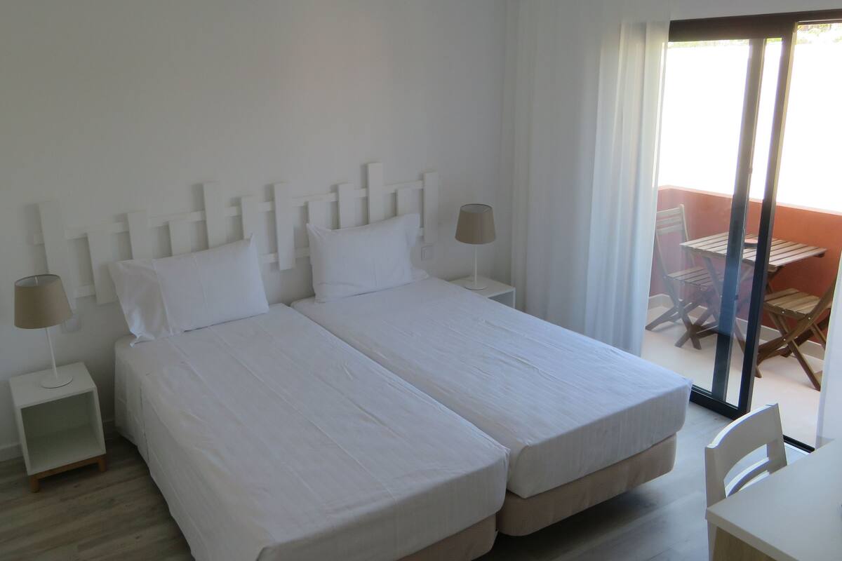 Uma cama de casal com lençóis brancos, duas mesas de cabeceira com abajures, e vista para uma varanda externa com mesa e cadeiras.