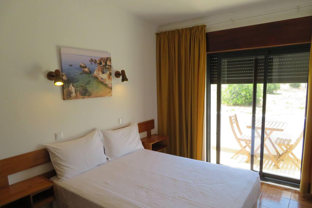 Quarto com cama, quadro, janela com cortinas e vista para o exterior.