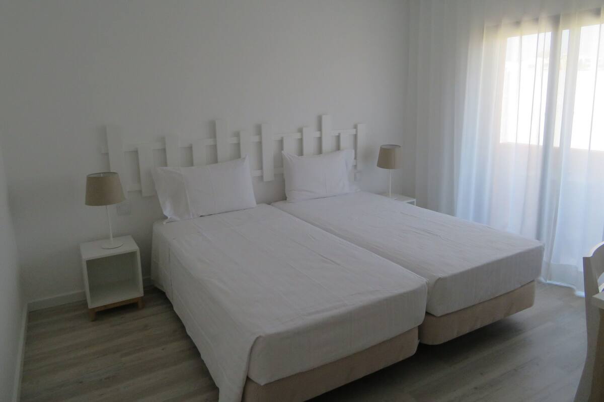 Uma cama de casal com cabeceira branca e duas mesas de cabeceira com abajures.