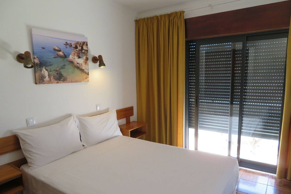 Uma cama com lençóis brancos, cabeceira de madeira, mesa de cabeceira, luminária na parede, um quadro e uma janela com persianas.