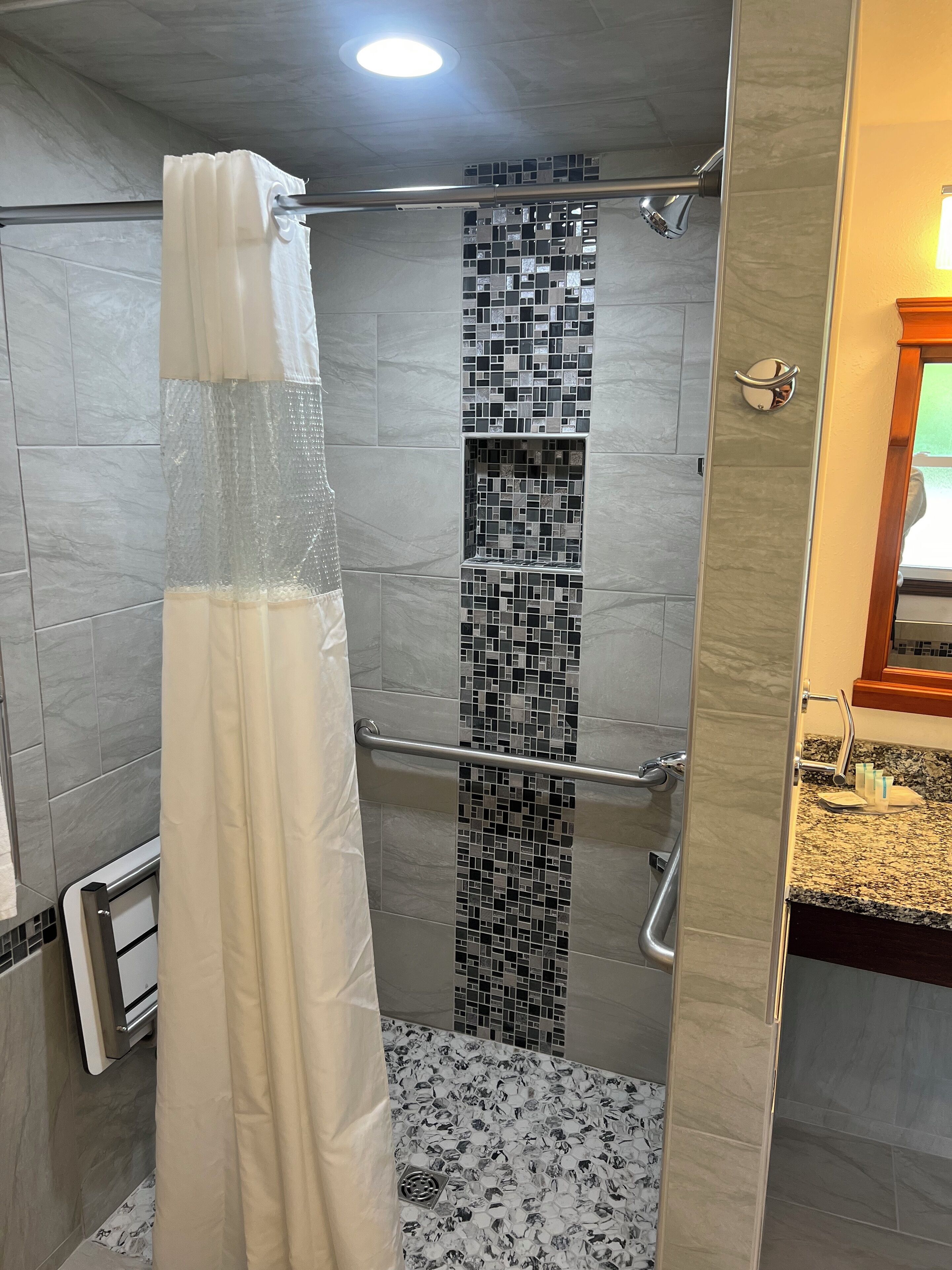 Área de chuveiro com parede revestida de azulejos, cortina de chuveiro, barras de apoio e um pequeno lavatório com produtos de higiene pessoal.