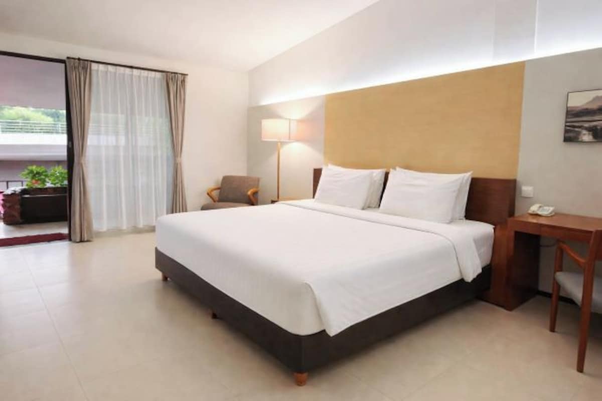 Quarto de hotel com uma cama grande, uma mesinha de cabeceira com abajur, uma cadeira e vista para uma varanda.
