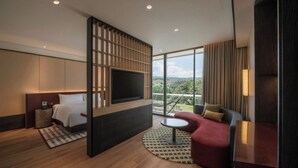 Quarto de hotel moderno com uma cama grande, uma televisão fixada em um painel de madeira, um sofá vermelho e uma varanda com vista para o campo.