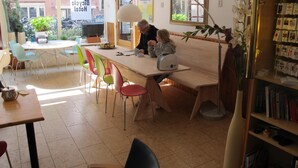 Um café acolhedor com uma mesa e cadeiras de madeira, uma estante de livros e uma janela grande com vista para a rua.