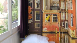 Quarto de hotel com cama, janela com cortina e um mural de paisagem urbana na parede.