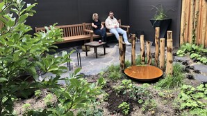 Uma área externa moderna com um banco de madeira e uma pequena mesa, cercada por vegetação e um cercado de madeira decorativo.