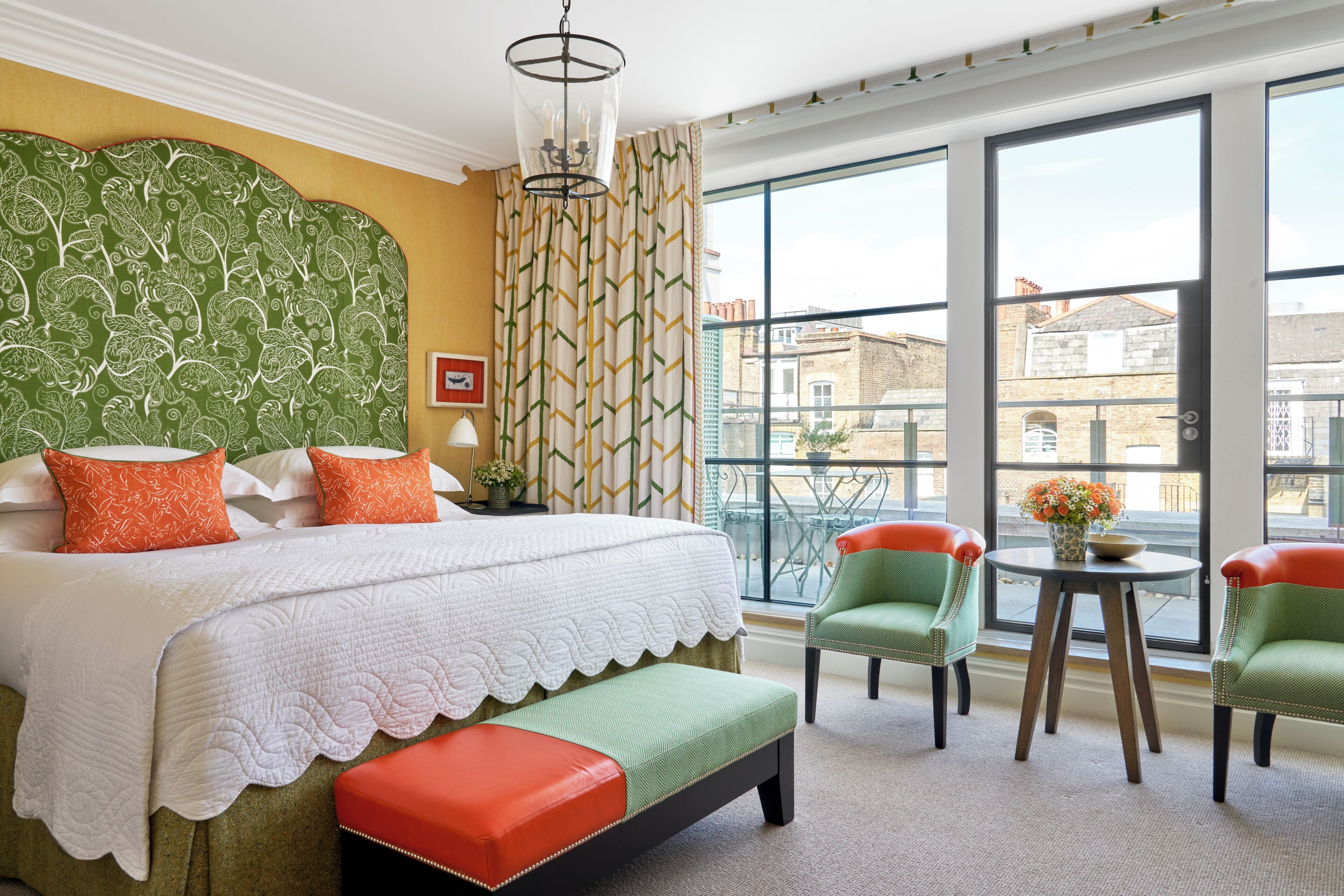 Quarto de hotel com uma cama grande, cadeiras verdes e laranja, uma mesa pequena e vista da cidade através de grandes janelas.