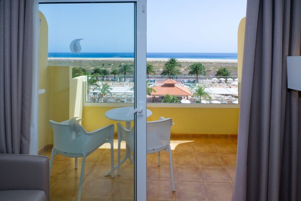 Varanda com duas cadeiras de vime brancas e uma mesa redonda pequena, com vista para uma praia com palmeiras e um edifício com telhado vermelho.