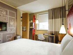 Quarto de hotel com cama, escrivaninha, cadeira, televisão, guarda-roupa e janela com cortinas.