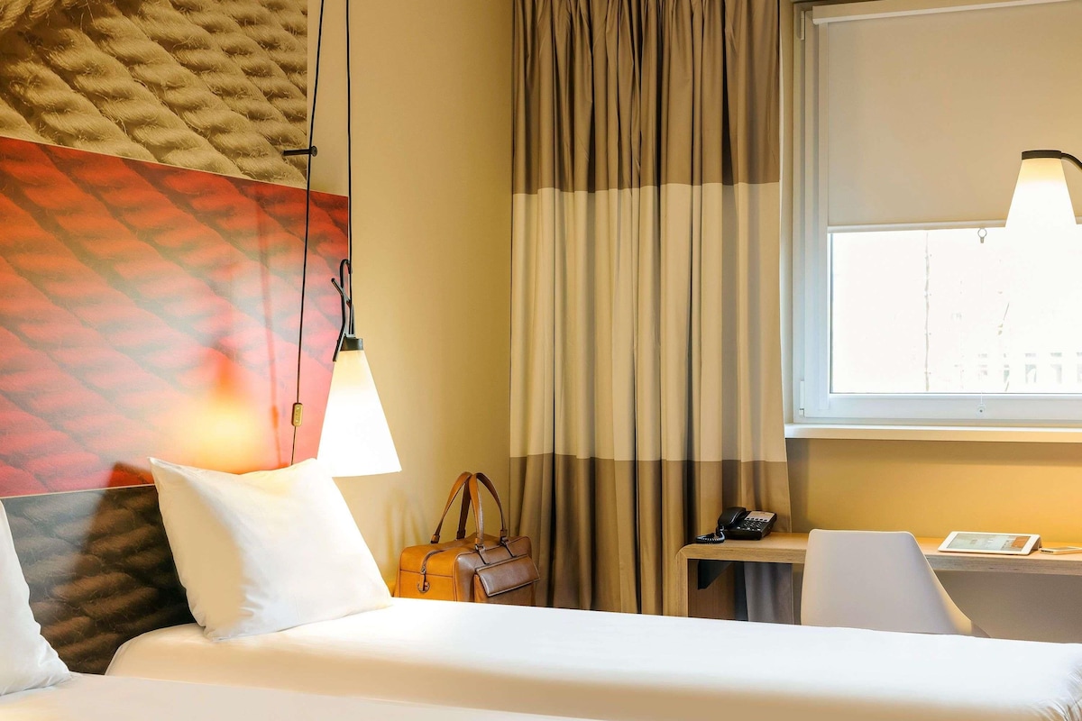 Quarto de hotel com cama, escrivaninha, cadeira, luminária, bolsa de mão e janela com cortinas.