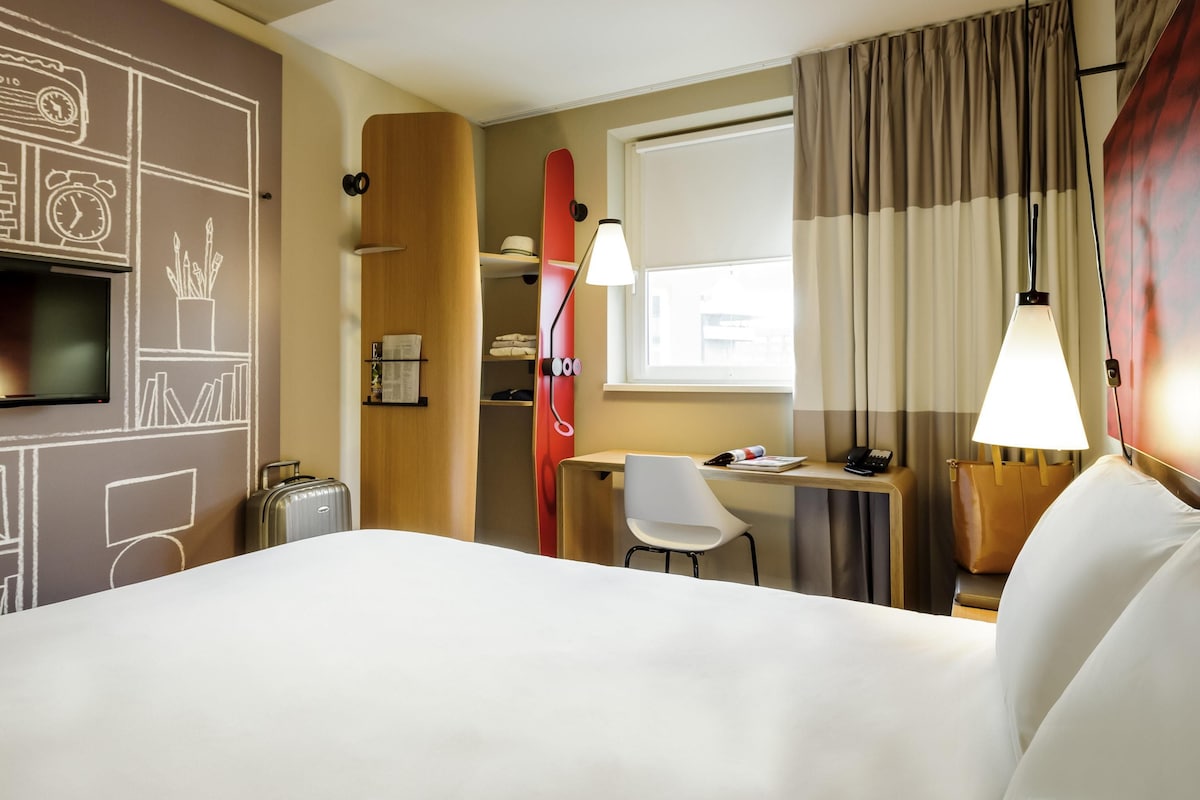 Quarto de hotel com cama, escrivaninha, cadeira, televisão, guarda-roupa e janela com cortinas.