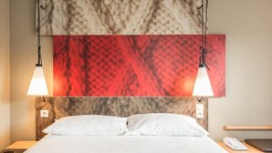 Quarto de hotel com cama, duas luminárias de cabeceira, mesa de cabeceira com telefone e uma pintura abstrata vermelha acima da cama.