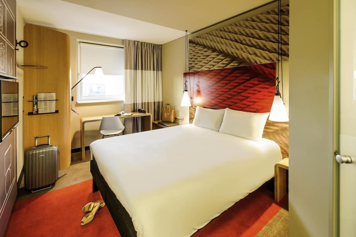 Quarto de hotel com cama, escrivaninha, cadeira, mala e uma parede de destaque vermelha.