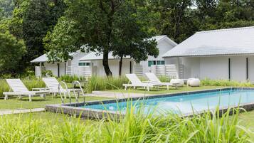 Uma área de piscina com espreguiçadeiras, uma piscina e edifícios brancos com venezianas verdes.
