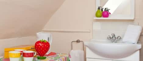 Um banheiro com uma pia branca, um espelho e uma mesa com toalha de mesa estampada com frutas. Há copos coloridos e uma decoração em formato de morango sobre a mesa.