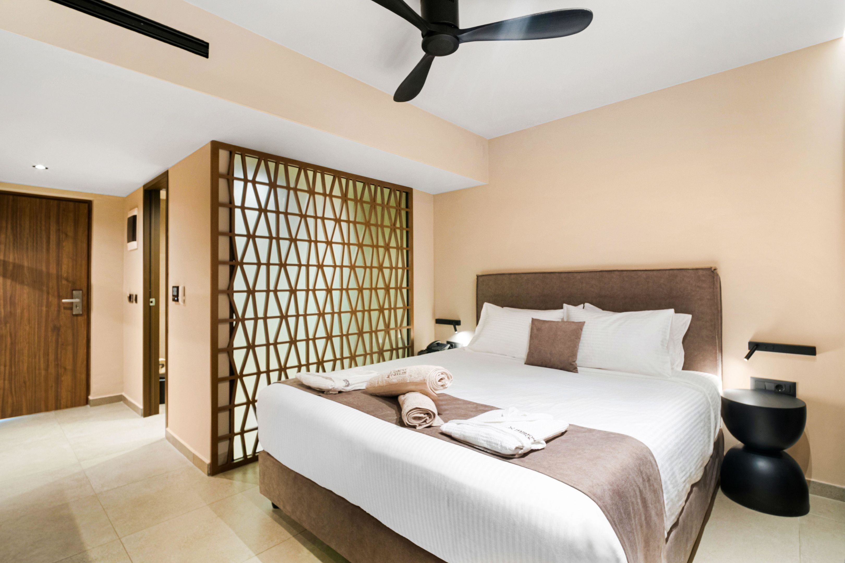 Quarto de hotel com uma cama grande, um ventilador de teto, uma divisória de madeira e uma mesa de cabeceira moderna.