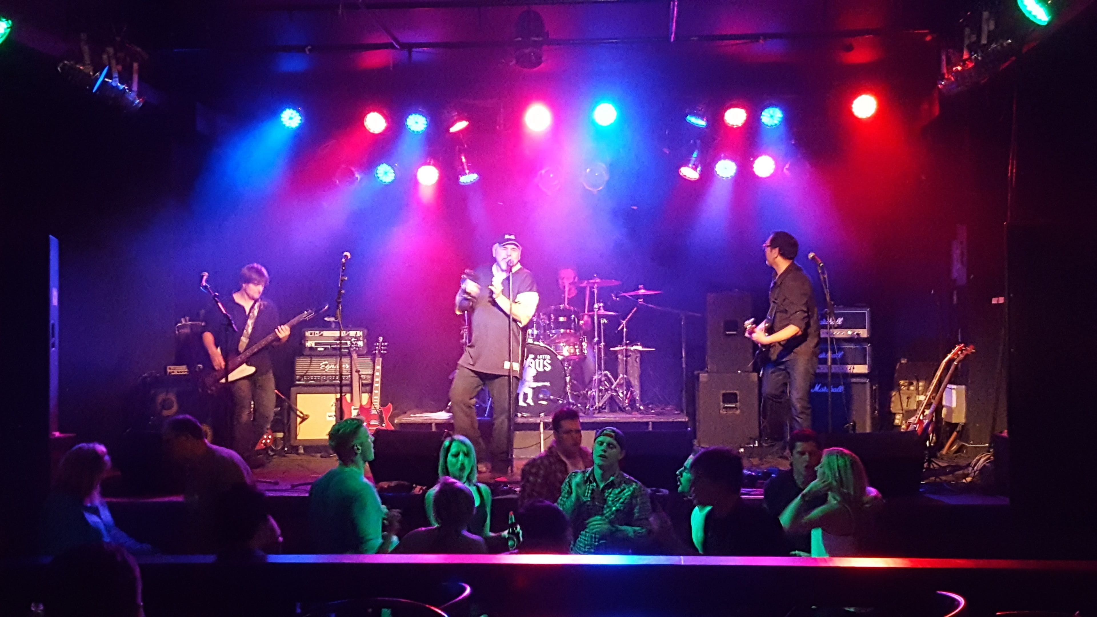 Uma banda se apresentando em um palco, com bateria e amplificadores ao fundo.