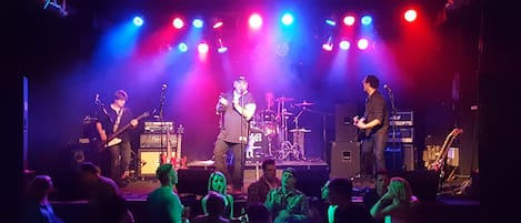 Uma banda se apresentando em um palco, com bateria e amplificadores ao fundo.