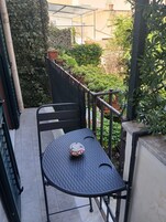 Uma pequena área de estar ao ar livre, com uma mesa e cadeiras, cercada por plantas em vasos e uma parede de pedra.