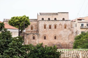 Um edifício histórico de pedra com janelas em arco e telhado de telhas, rodeado por vegetação e edifícios vizinhos.