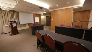 Uma sala de conferências com uma mesa comprida, cadeiras vermelhas, um pódio, uma tela e armários de madeira.