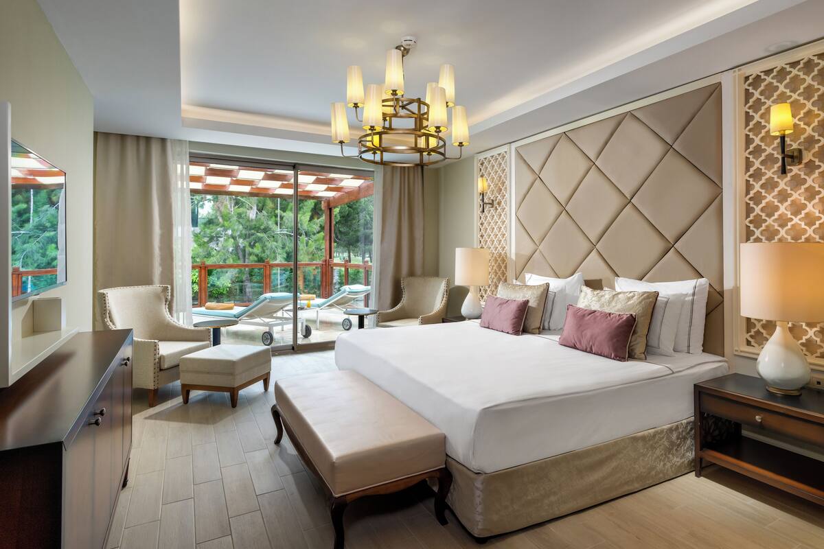 Um quarto espaçoso com uma cama grande, um espaço de estar com uma poltrona e um banco, uma pequena mesa, e vista para o exterior por uma janela ampla.