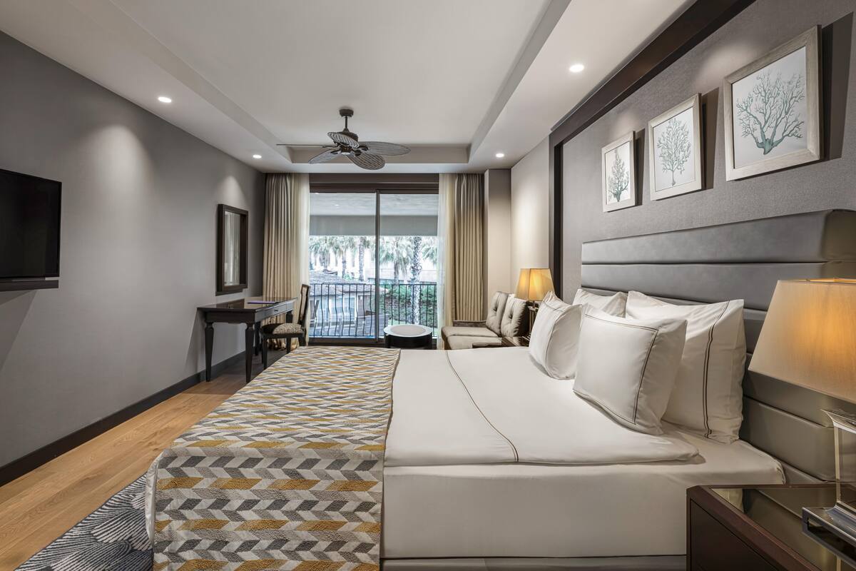 Quarto de hotel moderno com uma cama grande, uma mesa, uma cadeira e uma varanda com vista para palmeiras.