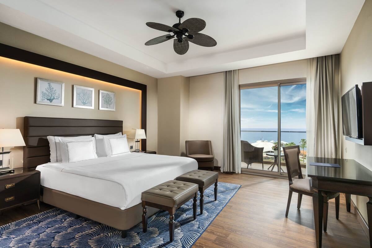 Quarto de hotel com uma cama grande, uma mesa, uma cadeira, um ventilador de teto e vista para o mar.