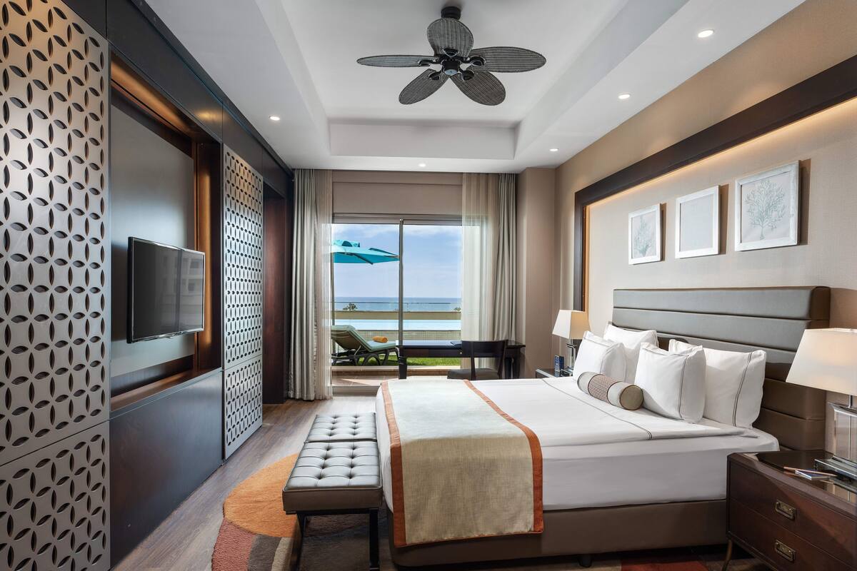 Quarto de hotel com cama, televisão, ventoinha no teto, vista para o oceano e varanda com uma cadeira.