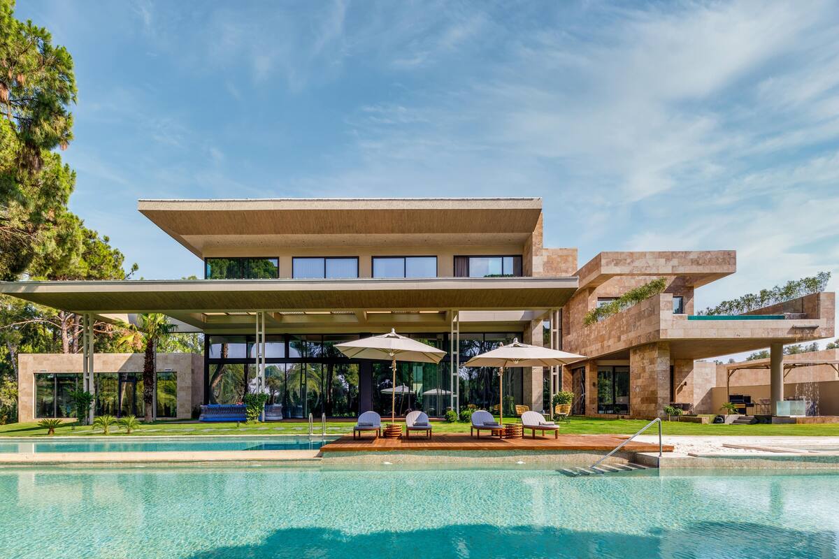 Uma área moderna à beira da piscina com espreguiçadeiras e guarda-sóis, piscina cristalina e um edifício contemporâneo com grandes janelas.