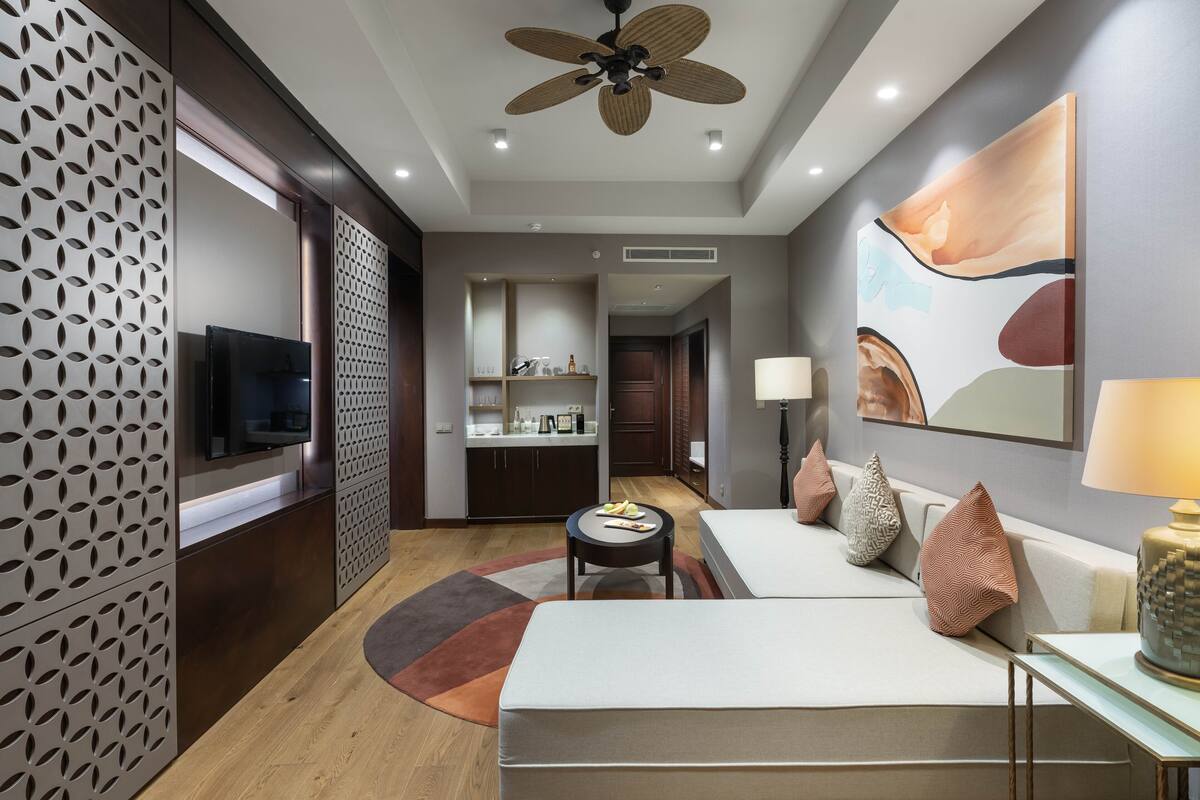 Quarto de hotel moderno com TV de tela plana, ventilador de teto, sofá com almofadas, pequena mesa com luminária e área de cozinha ao fundo.
