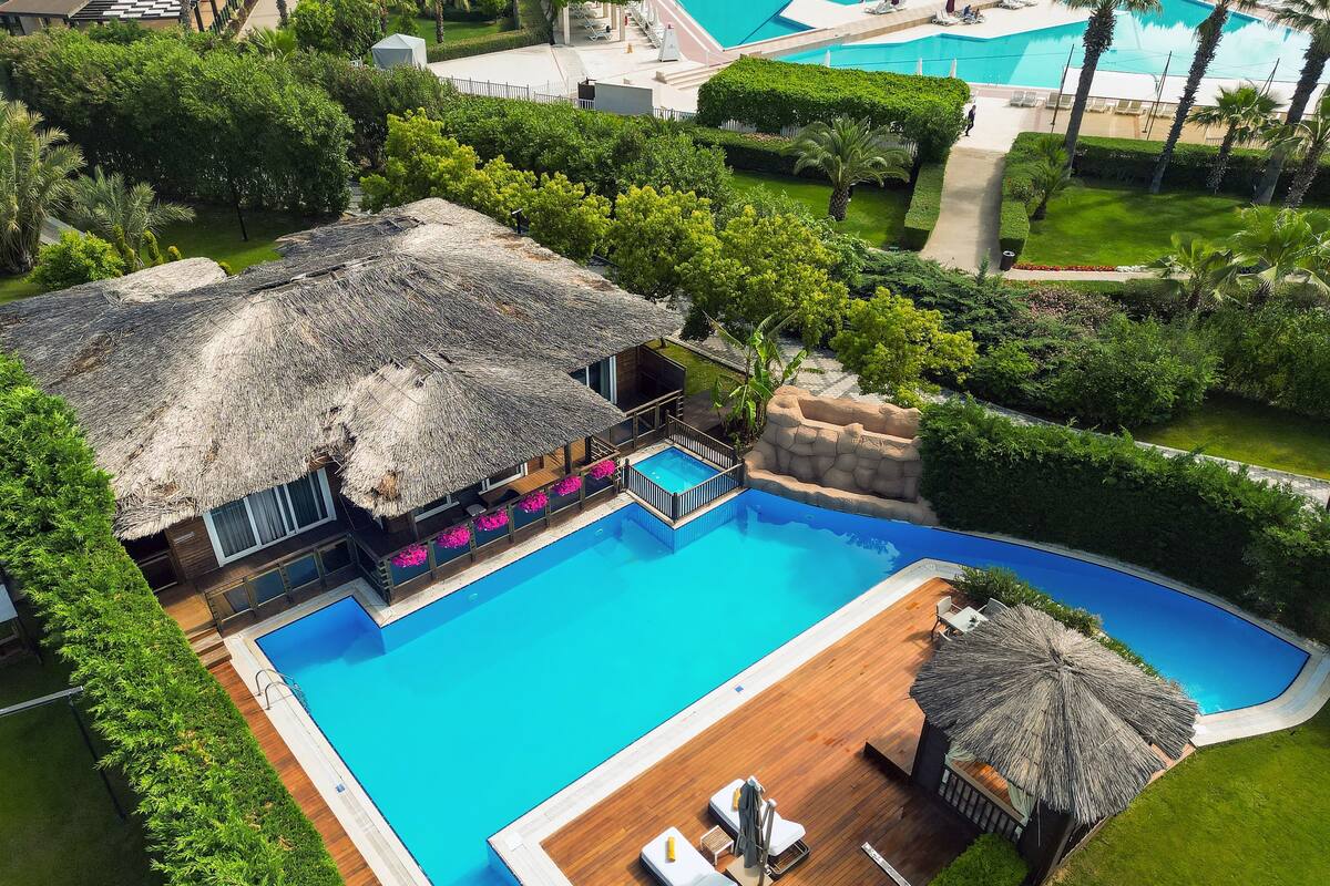 Um resort com uma grande piscina, uma estrutura com telhado de palha e vegetação exuberante.