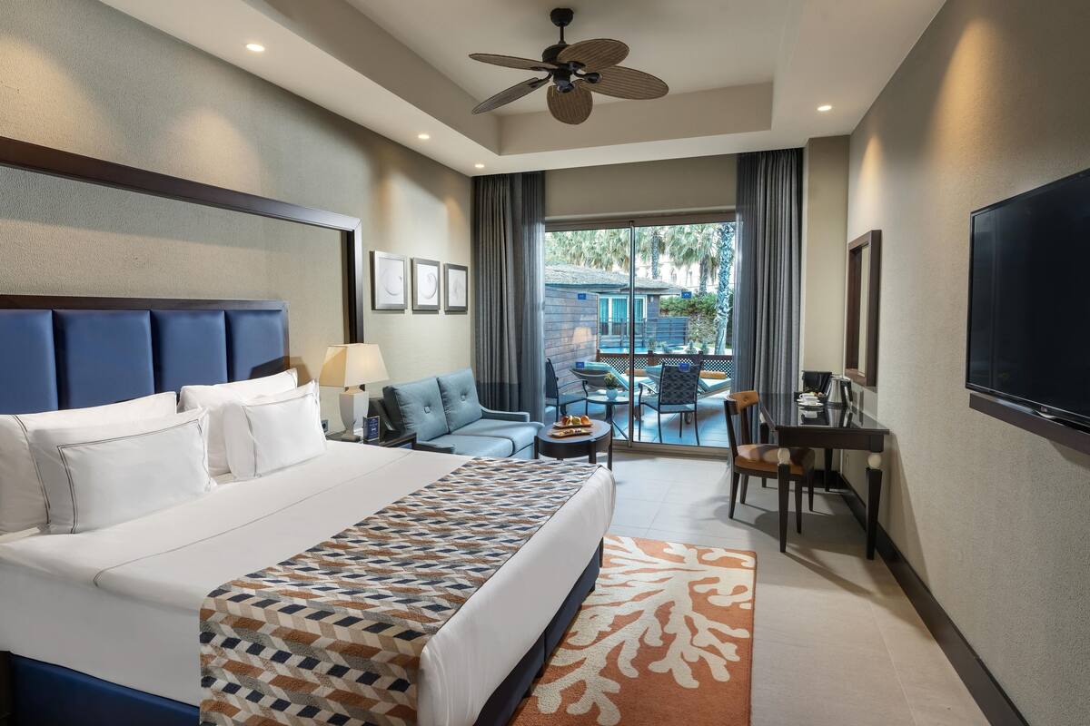 Quarto de hotel com uma cama grande, uma mesa, uma cadeira, uma televisão e uma varanda com vista para o exterior.
