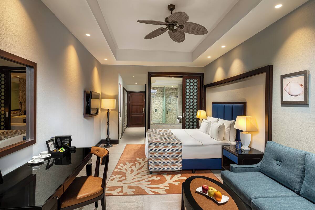Quarto de hotel com cama, sofá, escrivaninha com cadeira, mesinha com frutas e ventilador de teto.
