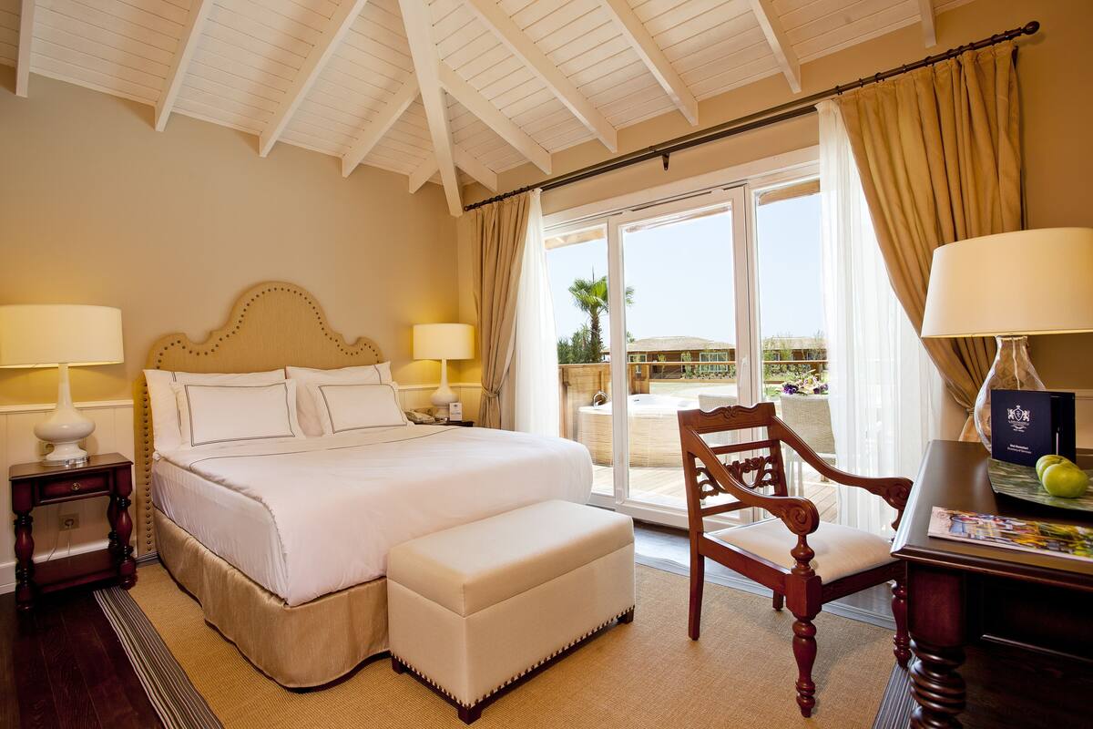 Quarto de hotel com uma cama grande, uma cadeira de madeira, uma escrivaninha com abajur e vista para o exterior.
