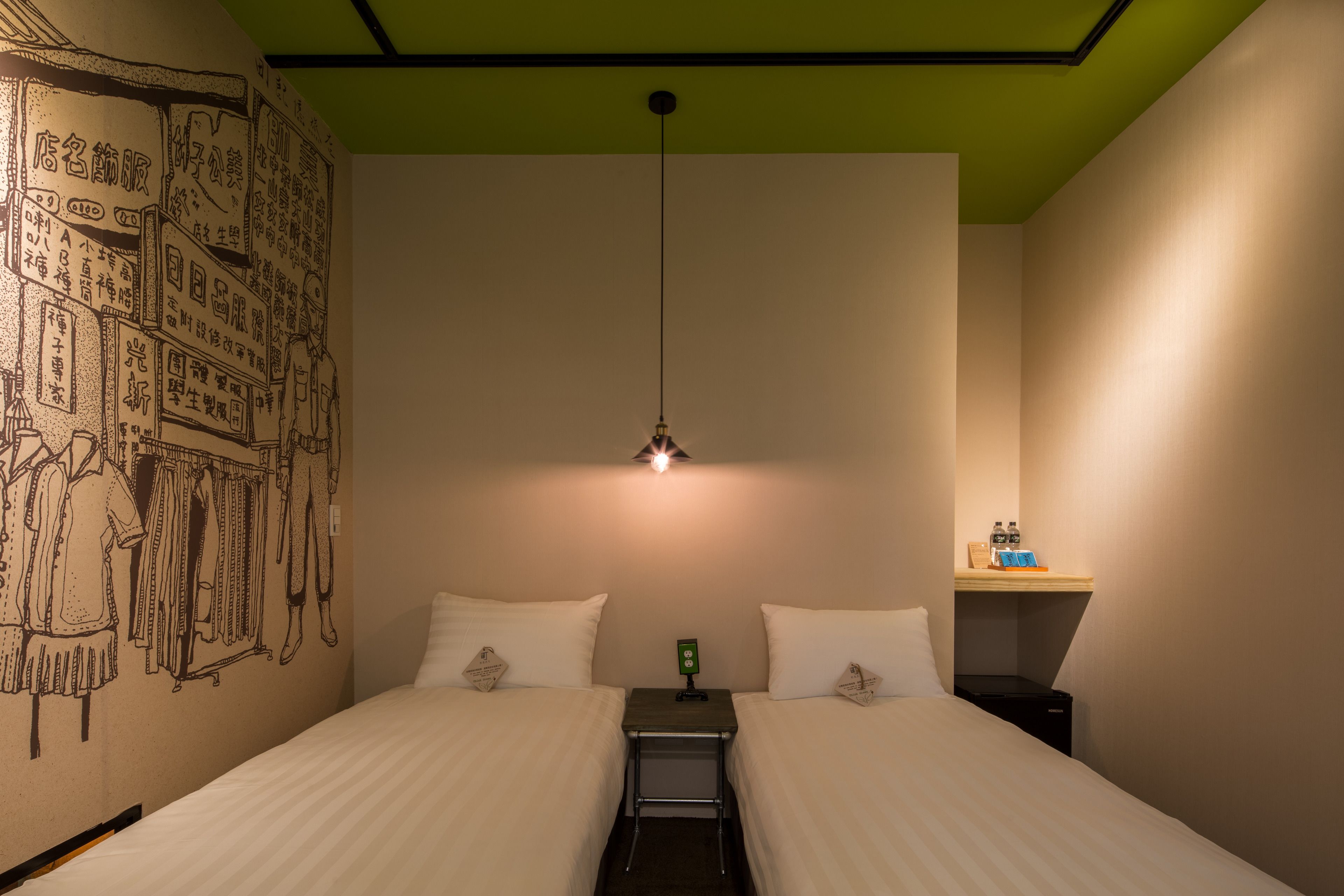 Quarto de hotel com duas camas, teto verde, parede com desenho em preto e branco, mesa de cabeceira com abajur e uma pequena prateleira com garrafas.