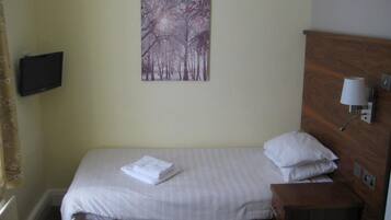 Uma cama bem arrumada com lençóis brancos e uma toalha dobrada, uma TV fixada na parede, um quadro com árvores e um abajur de cabeceira.