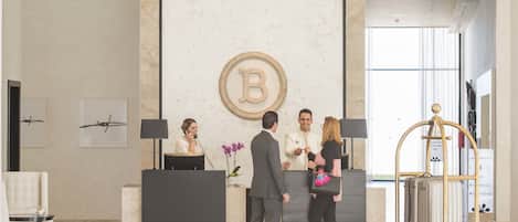 Uma área de recepção com balcão de mármore, um grande logotipo 'B', um balcão com computador, um carrinho de malas dourado e uma área de estar com sofá branco e uma pequena mesa.