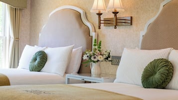 Quarto de hotel com duas camas, cada uma com travesseiro e mesa de cabeceira. Há um vaso com flores na mesinha de apoio.