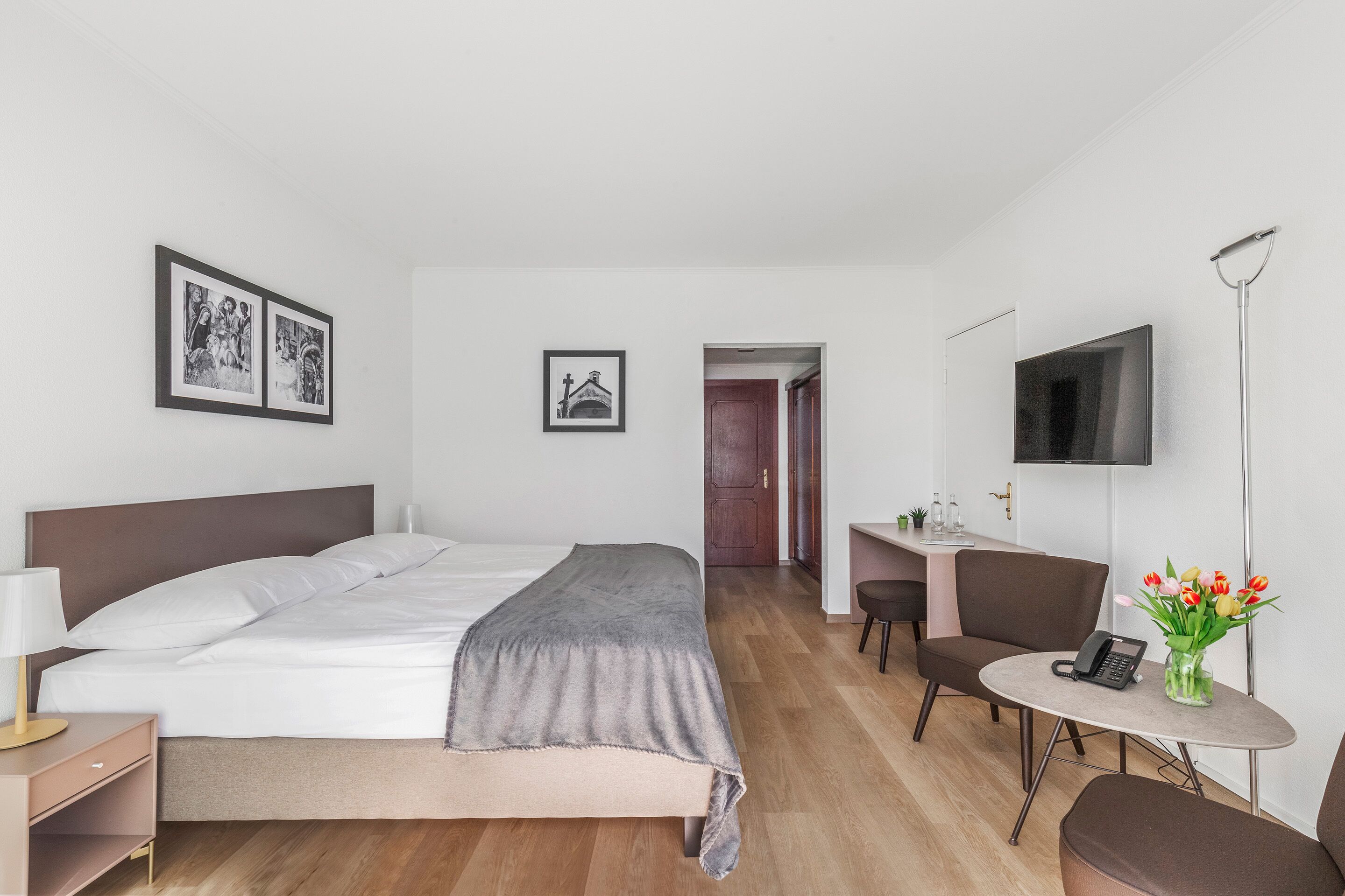 Quarto de hotel moderno com cama, mesa de cabeceira, abajur, escrivaninha, cadeira e televisão.