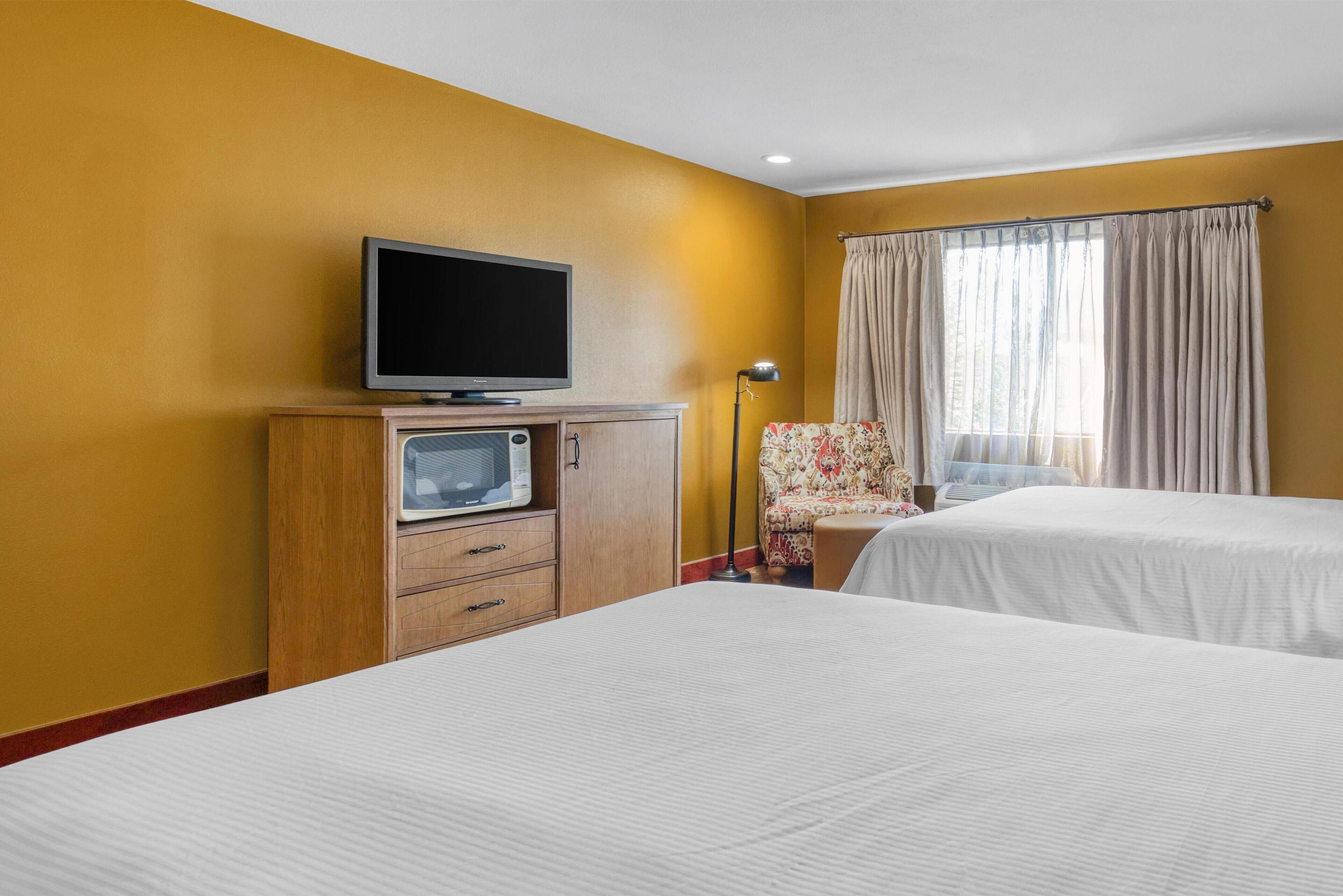 Quarto de hotel com duas camas, uma televisão sobre um armário de madeira, micro-ondas e uma cadeira com estofamento floral.
