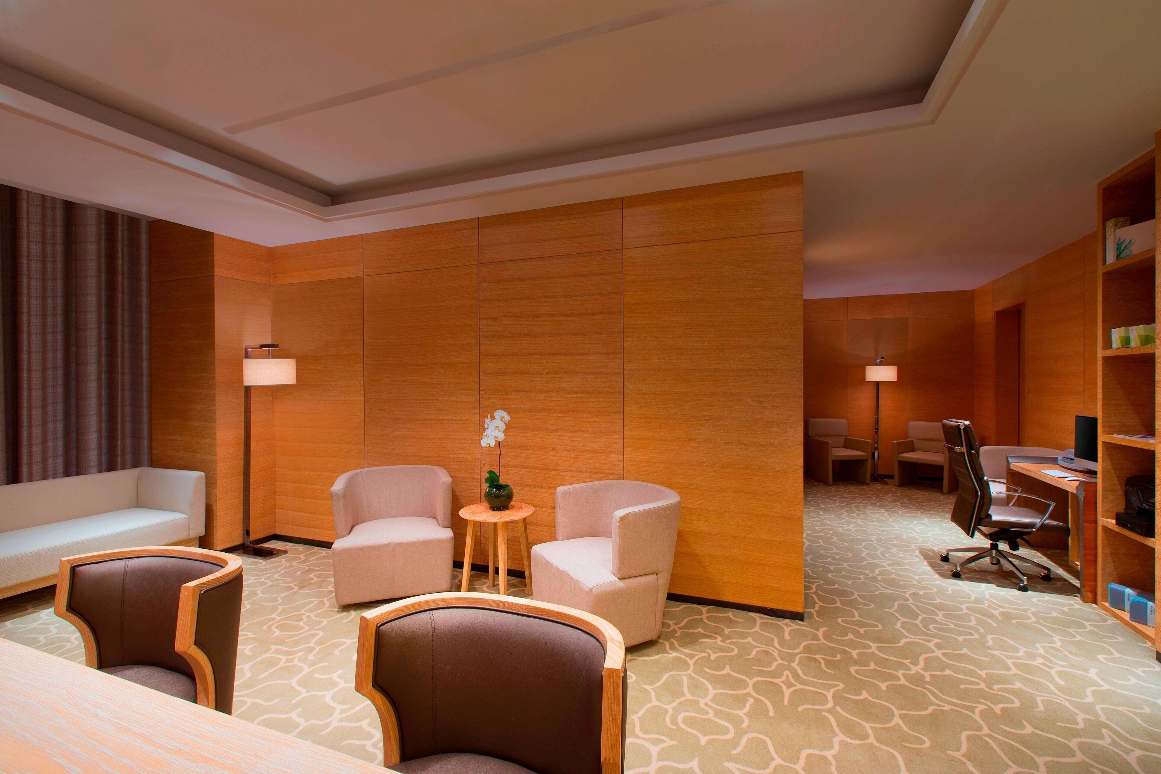 Quarto de hotel com sofá, duas poltronas, uma mesa pequena com abajur, escrivaninha com computador e estante de livros.