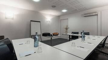 Uma sala de conferências com mesas brancas, cadeiras pretas, um quadro branco, uma pequena mesa com uma cesta e um aparelho de ar condicionado na parede.