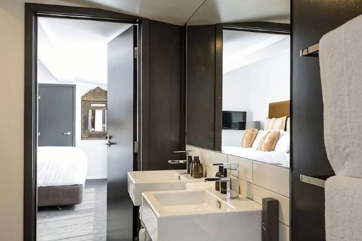 Um banheiro de hotel moderno com um espelho grande, duas pias e uma cama visível por uma porta aberta.