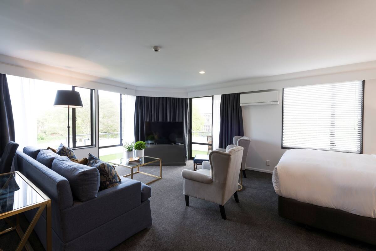 Quarto de hotel moderno com uma cama grande, um sofá, uma poltrona, televisão e vista para a varanda.