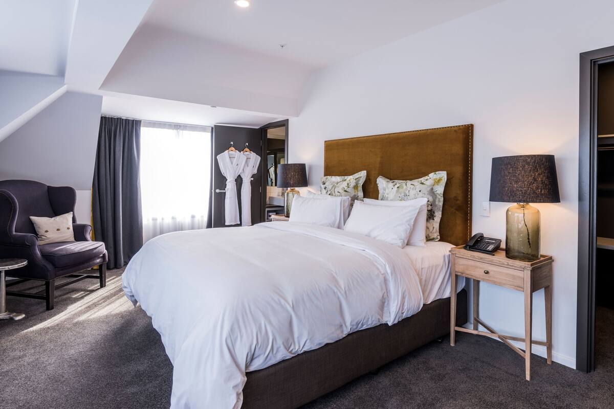 Quarto de hotel moderno com uma cama grande, uma cadeira, mesa de cabeceira com abajur e um espelho.