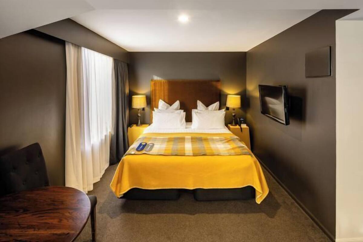 Quarto de hotel com uma cama, duas mesas de cabeceira com abajures, uma televisão na parede e uma cadeira.