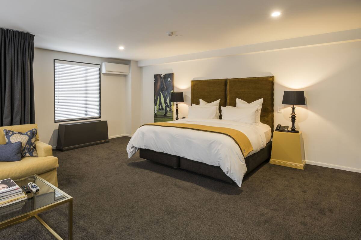 Quarto de hotel com cama, sofá, mesa de centro, um quadro e mesa de cabeceira com abajur.