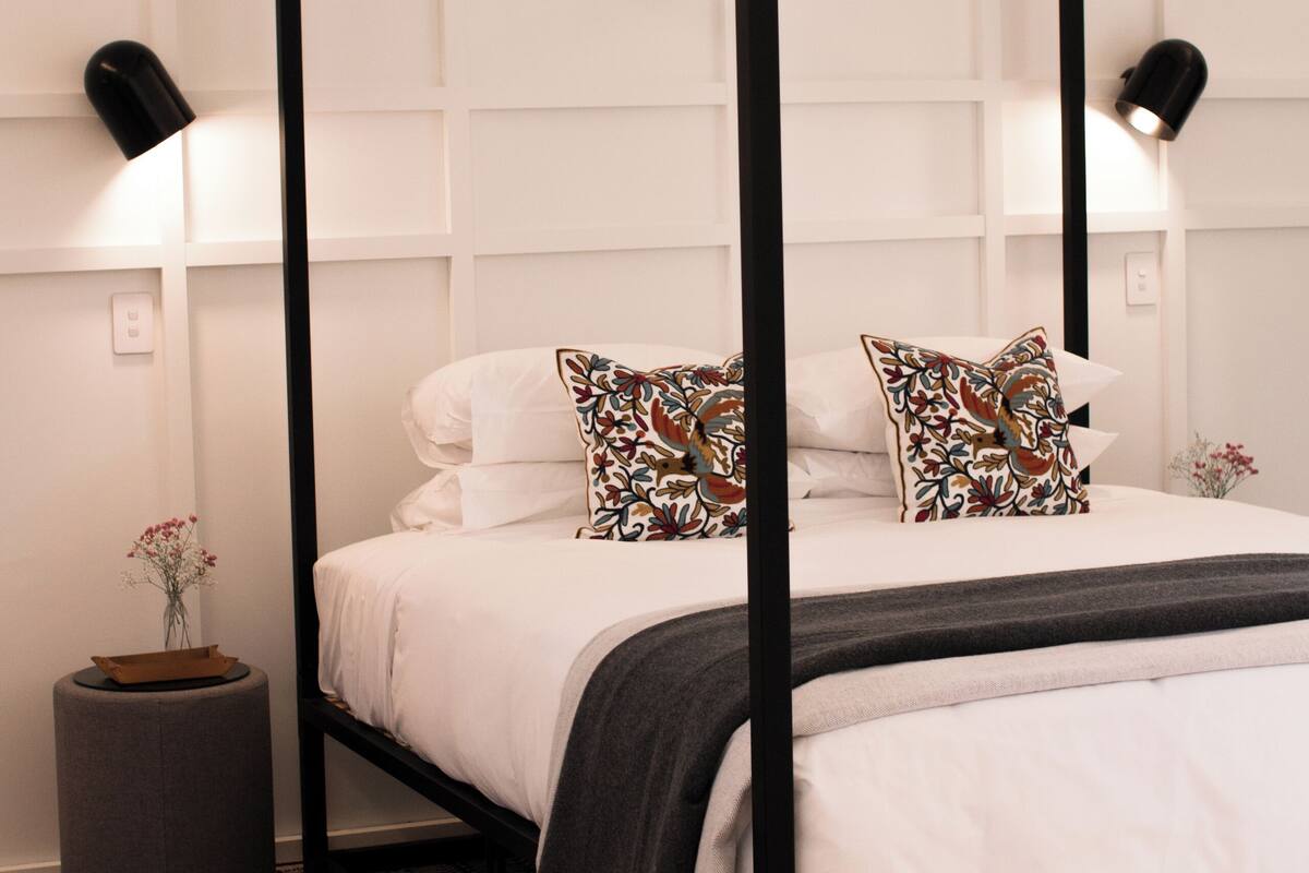 Uma cama de dossel com estrutura preta, roupa de cama branca e almofadas estampadas. Há um banquinho redondo pequeno com um vaso de flores ao lado da cama.