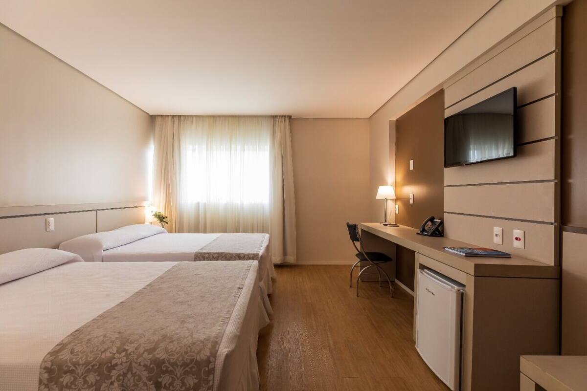 Quarto de hotel com duas camas, uma escrivaninha com cadeira, televisão na parede e um pequeno frigorífico.